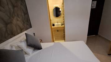 Quarto com cama, almofadas, mesa de cabeceira, espelho e banheiro visível através de um arco.