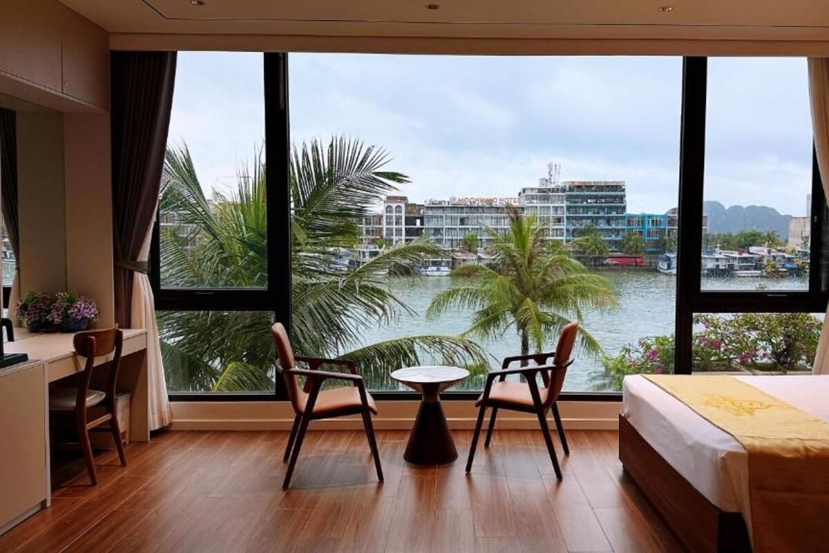 Quarto de hotel com uma grande janela, uma cama, uma escrivaninha e duas cadeiras.