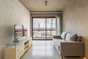 Uma sala de estar moderna com um sofá, uma TV em um móvel de vidro e uma janela grande com vista para a cidade.
