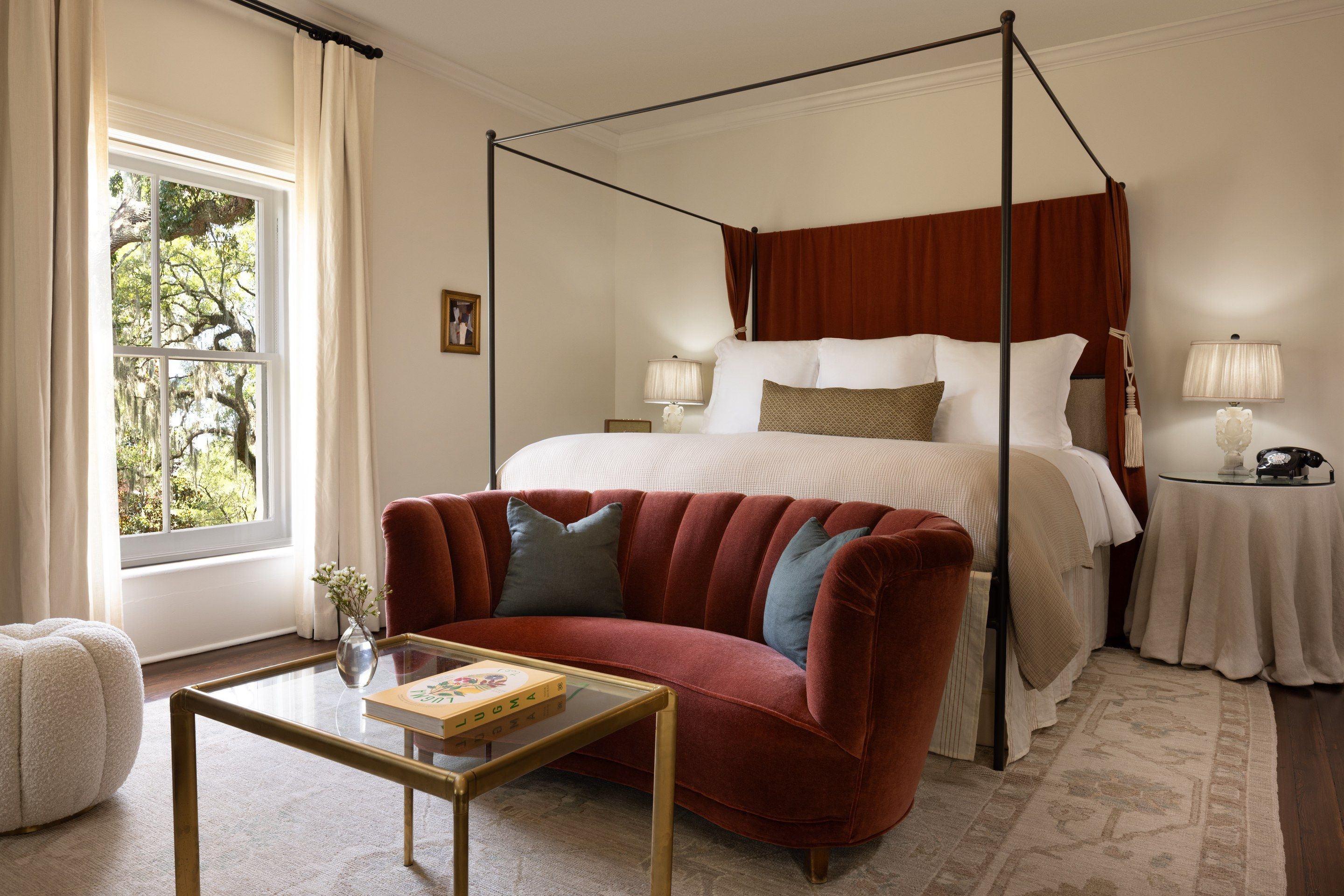 Uma cama com dossel e cortinas vermelhas, um sofá vermelho, uma mesinha de cabeceira com abajur e uma janela com cortinas.