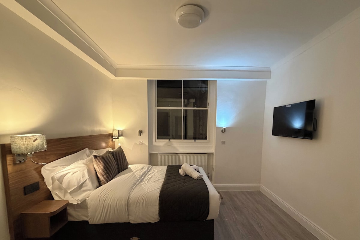 Quarto de hotel com cama, mesa de cabeceira, luminária, janela e televisão.
