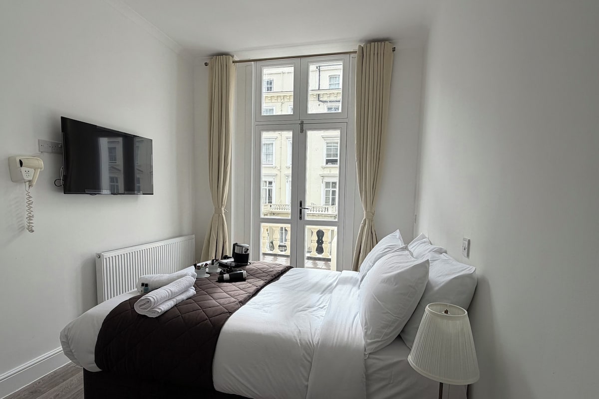 Quarto de hotel com uma cama, televisão na parede, janela com cortinas e abajur.