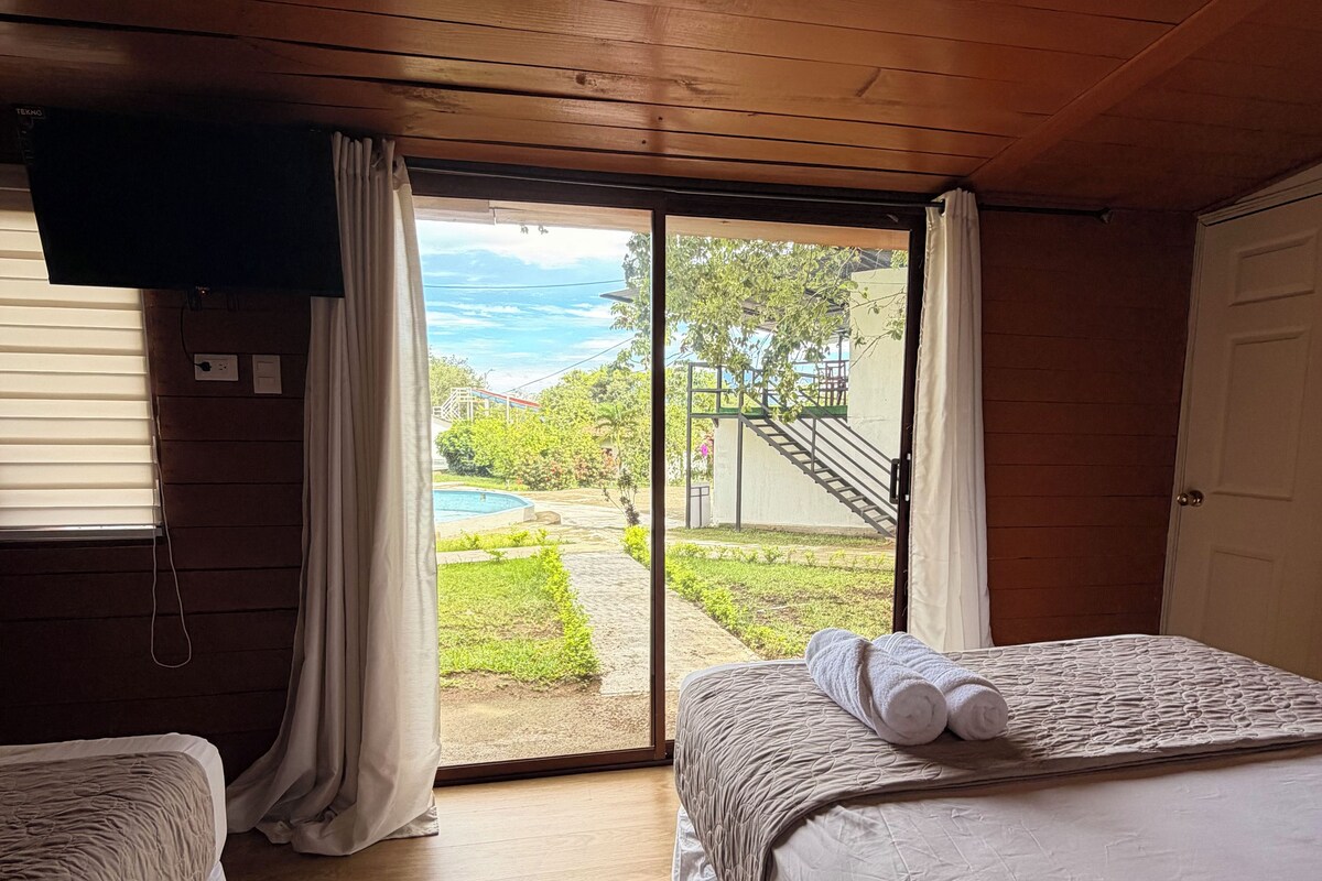 Quarto com duas camas, TV fixada na parede e porta de vidro deslizante para área externa com piscina.