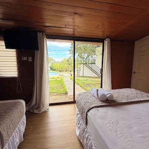 Quarto com duas camas, TV fixada na parede e porta de vidro deslizante para área externa com piscina.