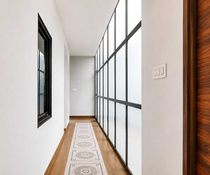 Um corredor com piso de madeira, paredes brancas e uma janela com moldura preta no lado esquerdo.