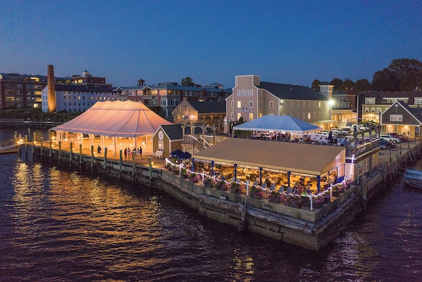 Restaurante à beira-mar com mesas ao ar livre, área coberta e um cais que se estende sobre a água.