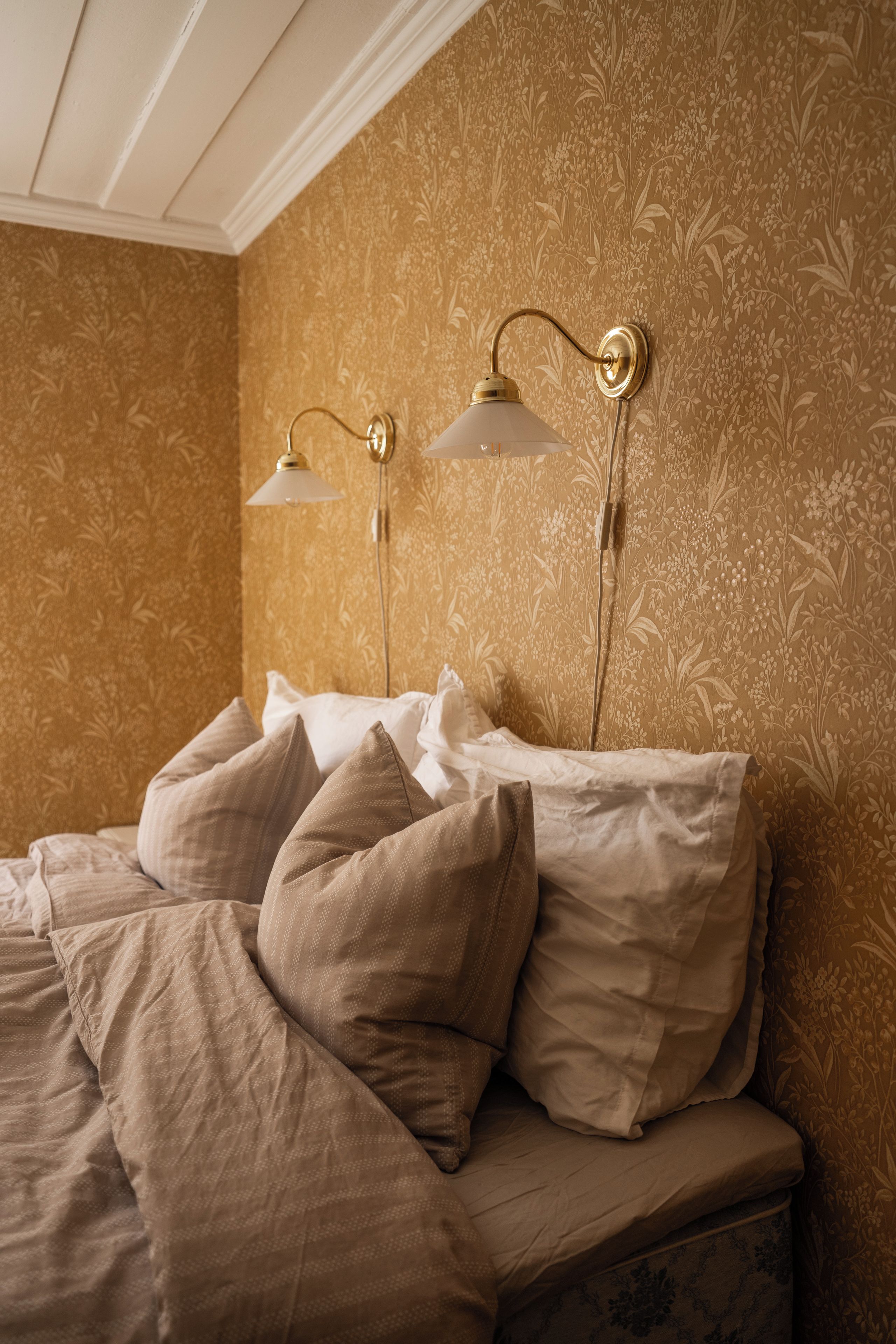 Quarto com cama, almofadas e duas luminárias fixadas na parede.