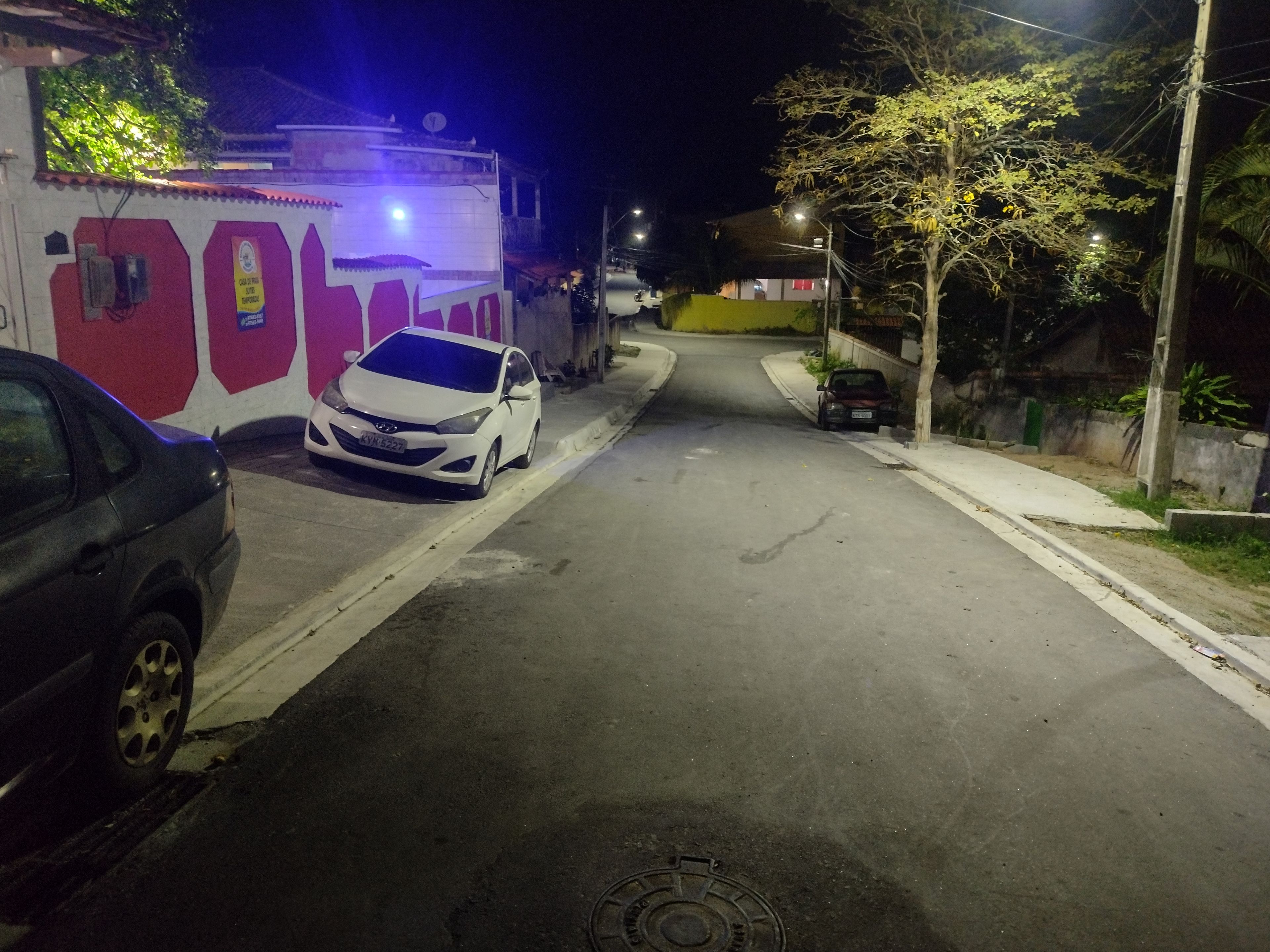 Cena noturna de rua com um carro branco estacionado na lateral, um prédio com fachada vermelha e branca, e uma árvore com flores amarelas.