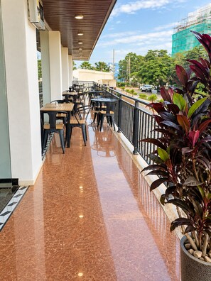 Uma área de estar coberta ao ar livre, com mesas e cadeiras, um vaso de planta e vista para a rua e vegetação.