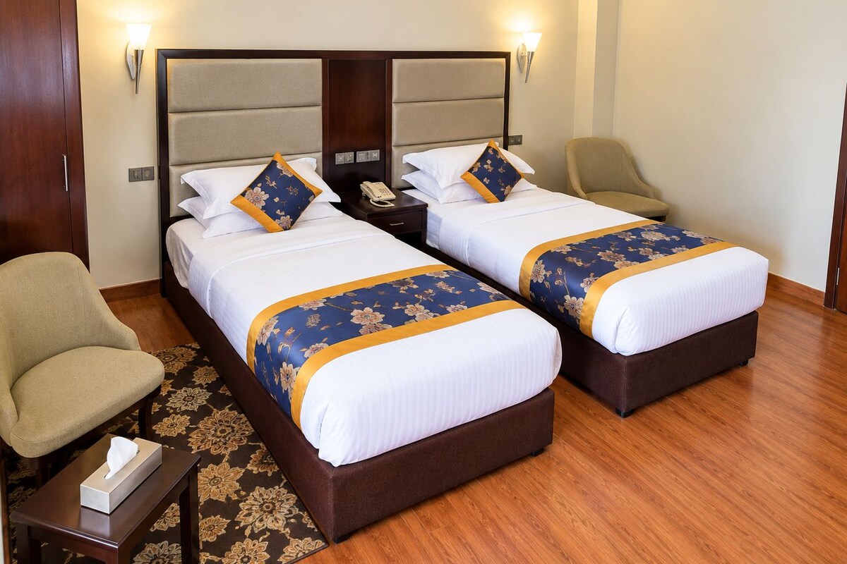 Quarto de hotel com duas camas, uma cadeira, mesa de cabeceira e piso coberto por carpete.