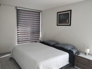 Quarto com cama, mesa de cabeceira, abajur, quadro na parede e janela com persianas.