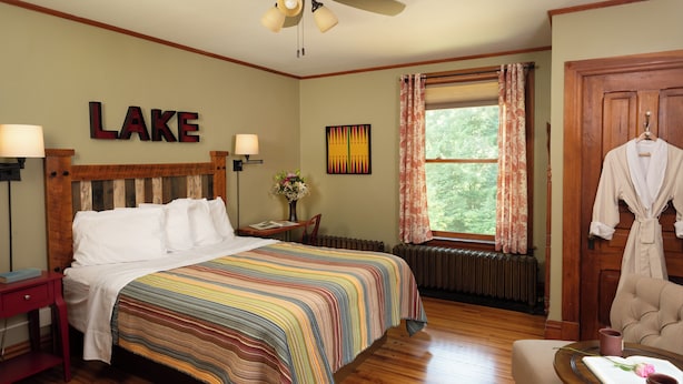 Quarto de hotel com cama, mesa de cabeceira, luminária para leitura, janela com cortinas, radiador, cadeira de madeira e roupão pendurado na porta.