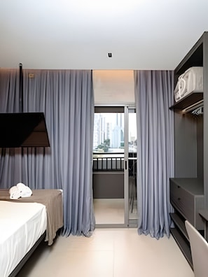 Quarto de hotel com cama, escrivaninha, cadeira e vista da cidade através de uma varanda.
