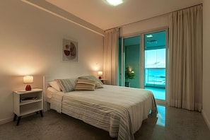 Quarto com cama, mesas de cabeceira, cortinas e vista para o mar.
