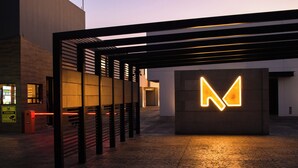 Entrada de um edifício moderno com um grande letreiro iluminado em forma de 'M', uma cobertura e calçada de tijolos.