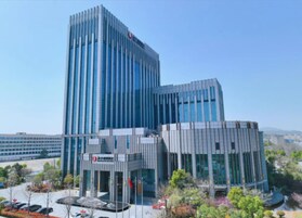 Um edifício moderno com design vertical marcante, entrada central e área de estacionamento com carros e vegetação.