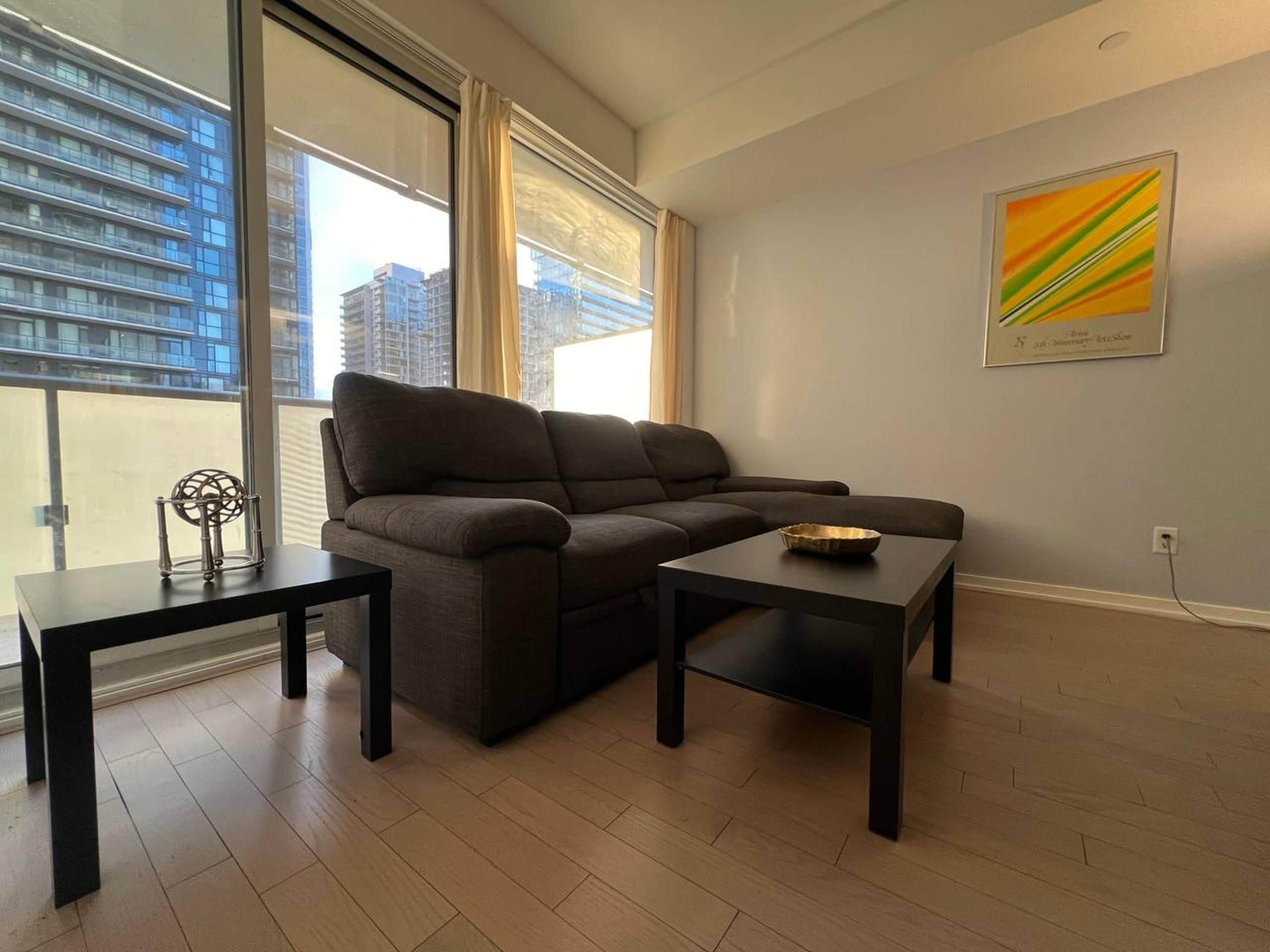 Uma sala de estar moderna com um sofá, duas mesas pretas e vista para a cidade através de grandes janelas.