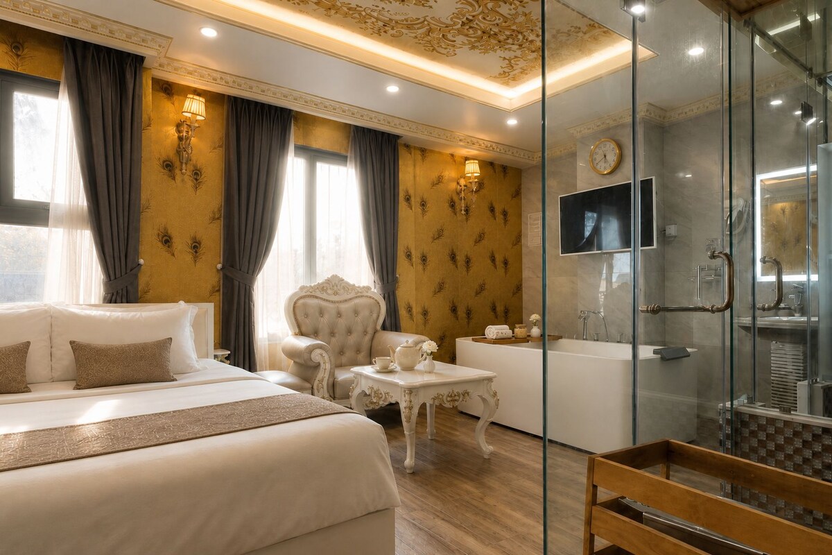 Quarto de hotel com uma cama grande, área de estar com uma poltrona e mesa, chuveiro em gabinete de vidro e TV montada na parede.