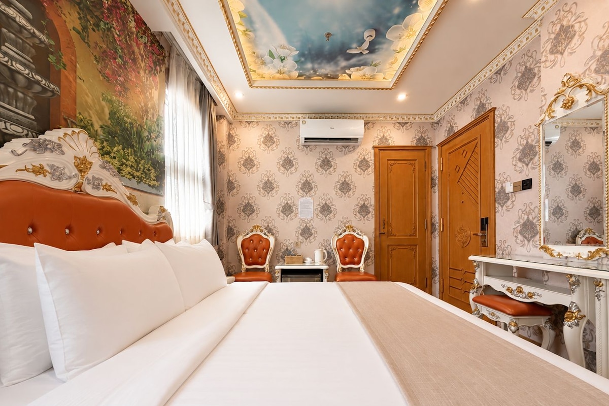 Quarto de hotel com uma cama grande, duas cadeiras, penteadeira com espelho e um mural no teto.