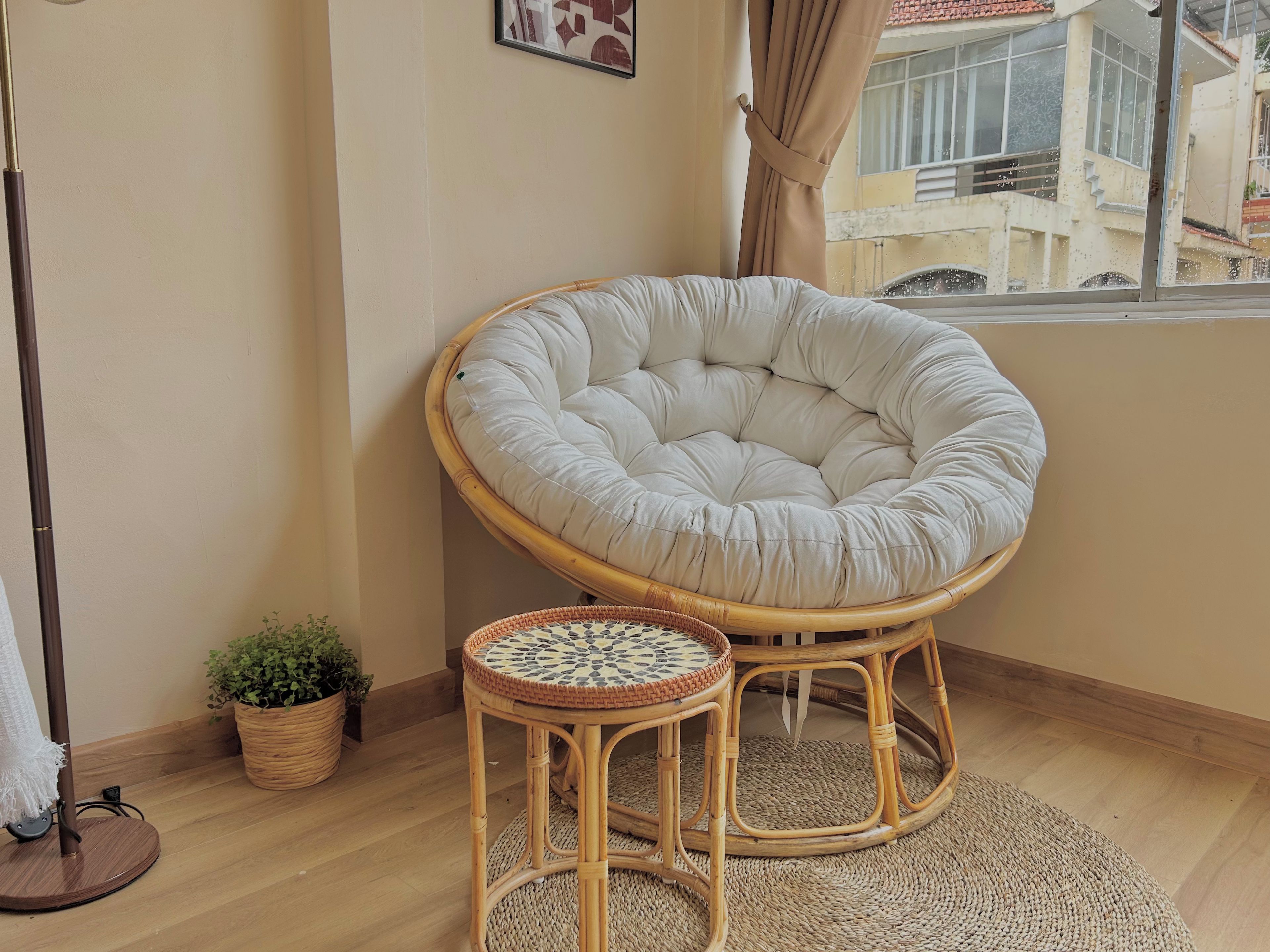 Quarto de canto com cadeira e banquinho de rattan, planta em vaso, e vista de prédios pela janela.