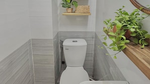 Uma casa de banho compacta com sanita, lavatório e espelho. Existem prateleiras com objetos decorativos e uma planta em vaso.