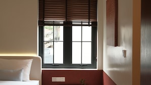Quarto com cama, janela com persianas, sofá, mesa pequena com um vaso e porta.