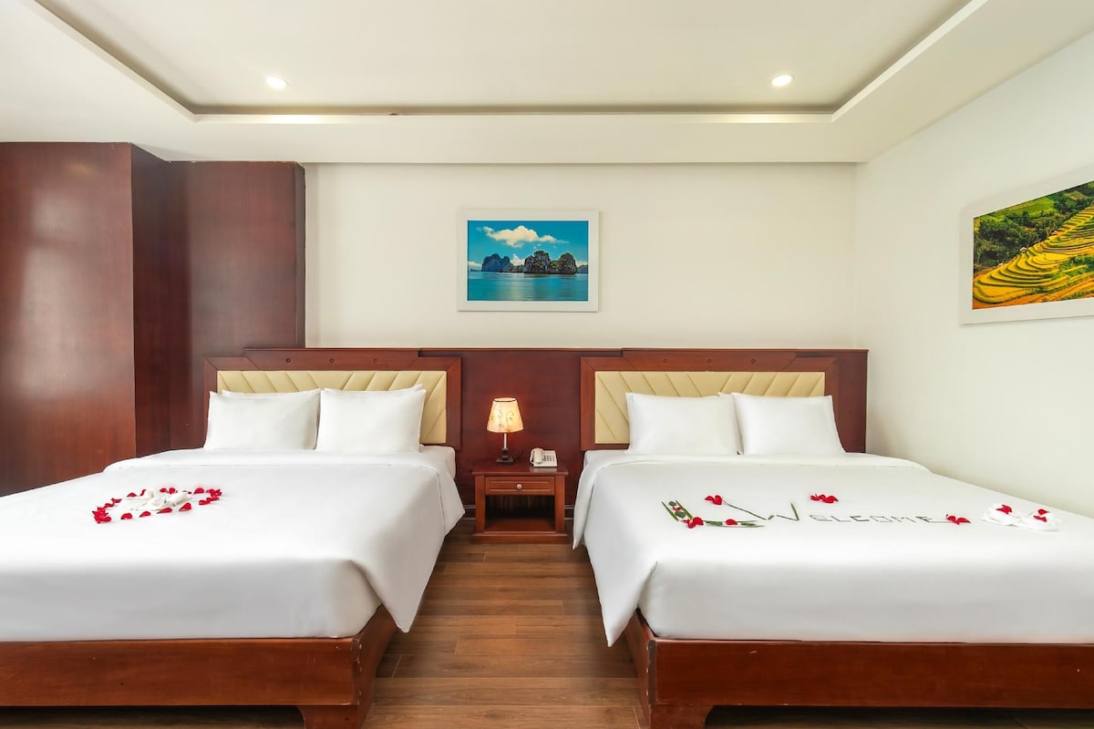 Quarto de hotel com duas camas, uma mesa de cabeceira com abajur e um quadro na parede.