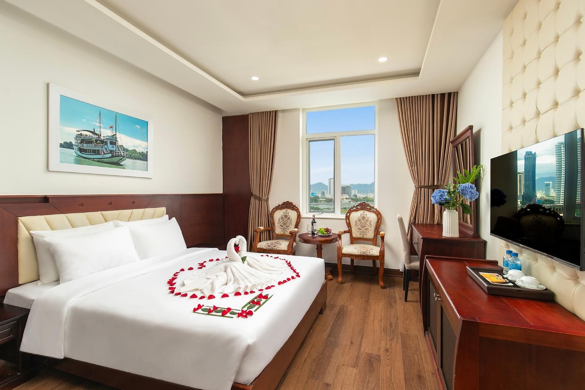 Quarto de hotel com uma cama grande, uma mesa, uma cadeira, uma televisão e uma janela com vista para a cidade.