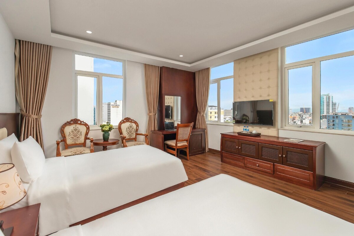 Quarto de hotel com duas camas, uma escrivaninha, televisão e vista para a cidade.