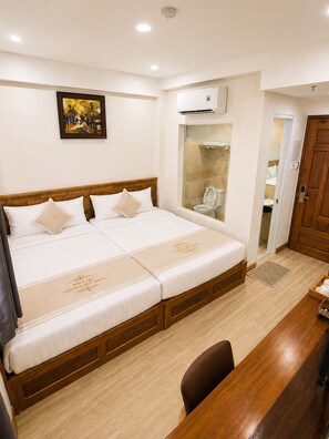 Quarto de hotel com cama de casal, escrivaninha de madeira, uma cadeira, guarda-roupa e banheiro visível através de uma porta de vidro.