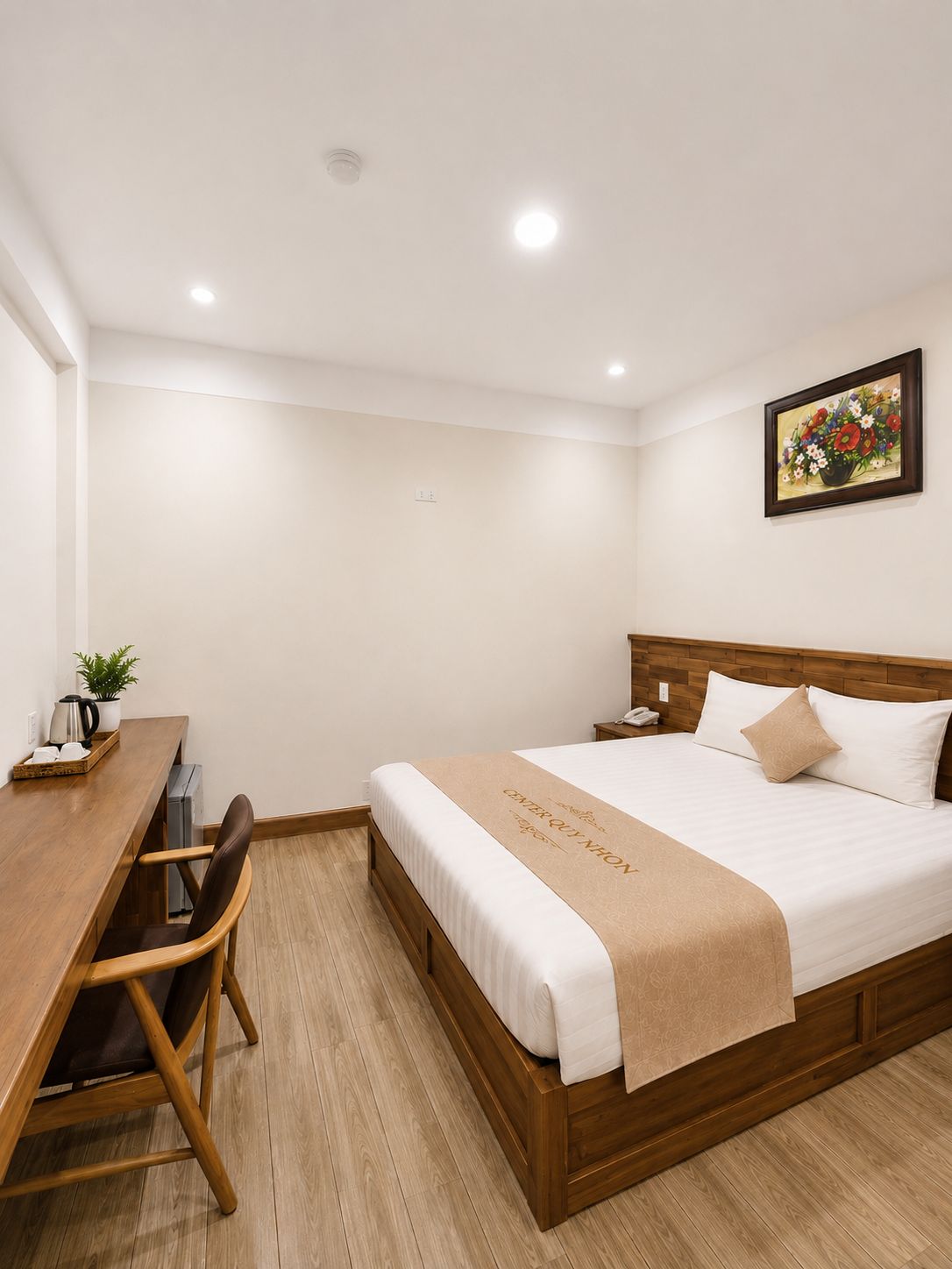 Quarto de hotel com cama de madeira, uma escrivaninha com cadeira, uma mesinha de centro com planta e um quadro na parede.