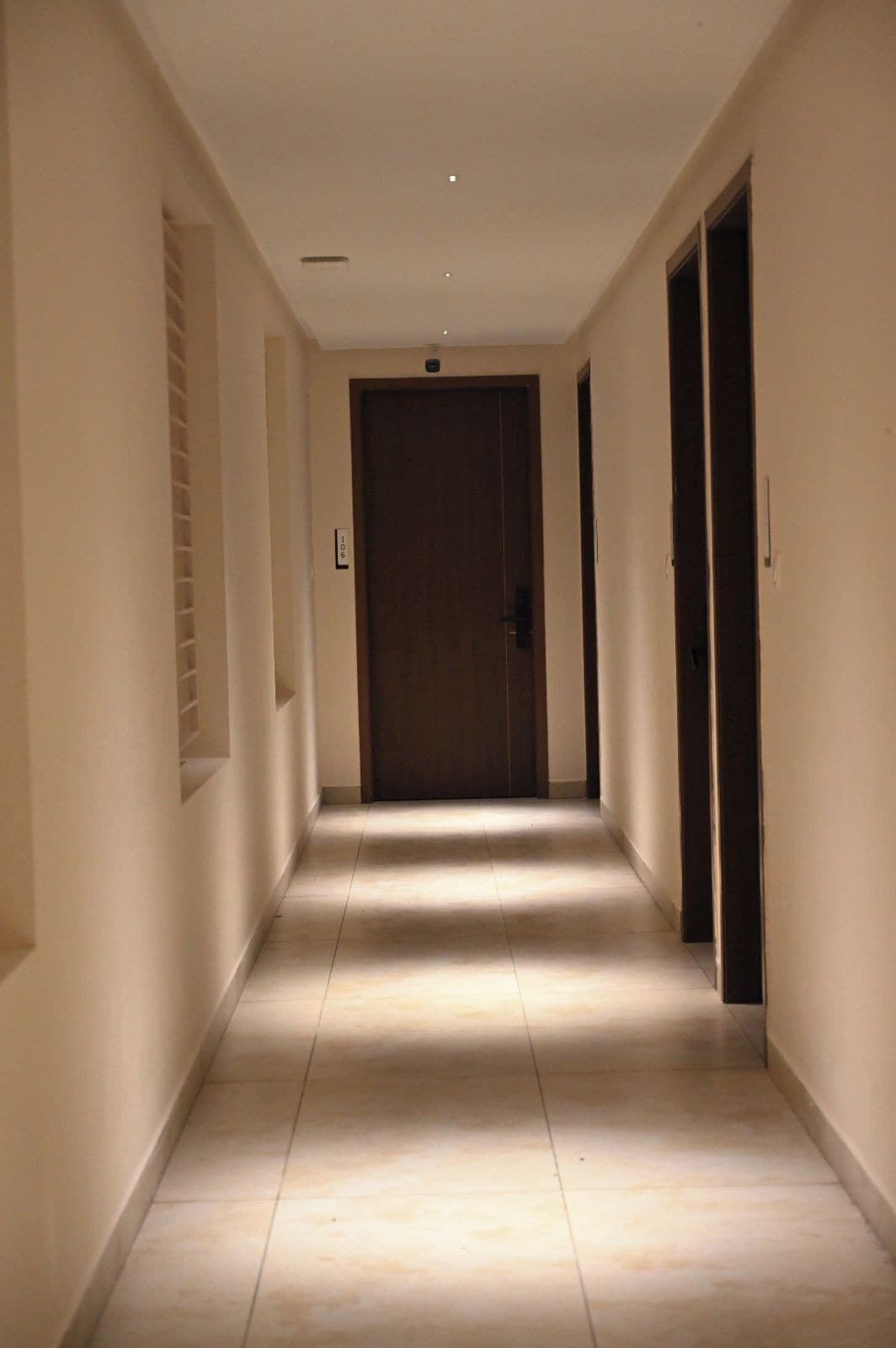 Um corredor com uma porta no fim, um luminário no teto e chão revestido com azulejos.