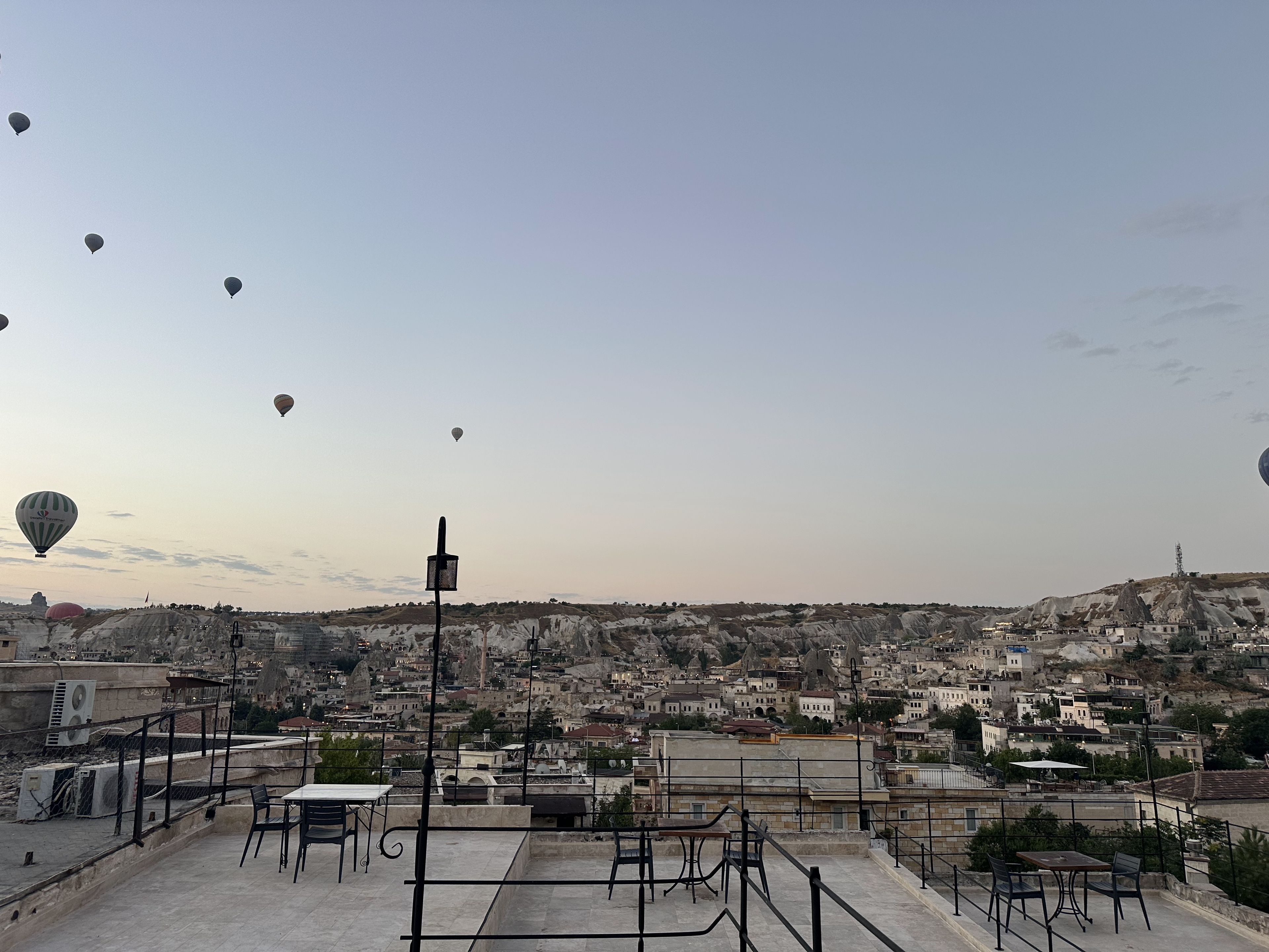 Terraço no telhado com mesas e cadeiras, vista para uma paisagem urbana com edifícios e balões de ar quente no céu.