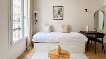 Quarto com cama, mesa de cabeceira, cadeira e janela com cortinas.