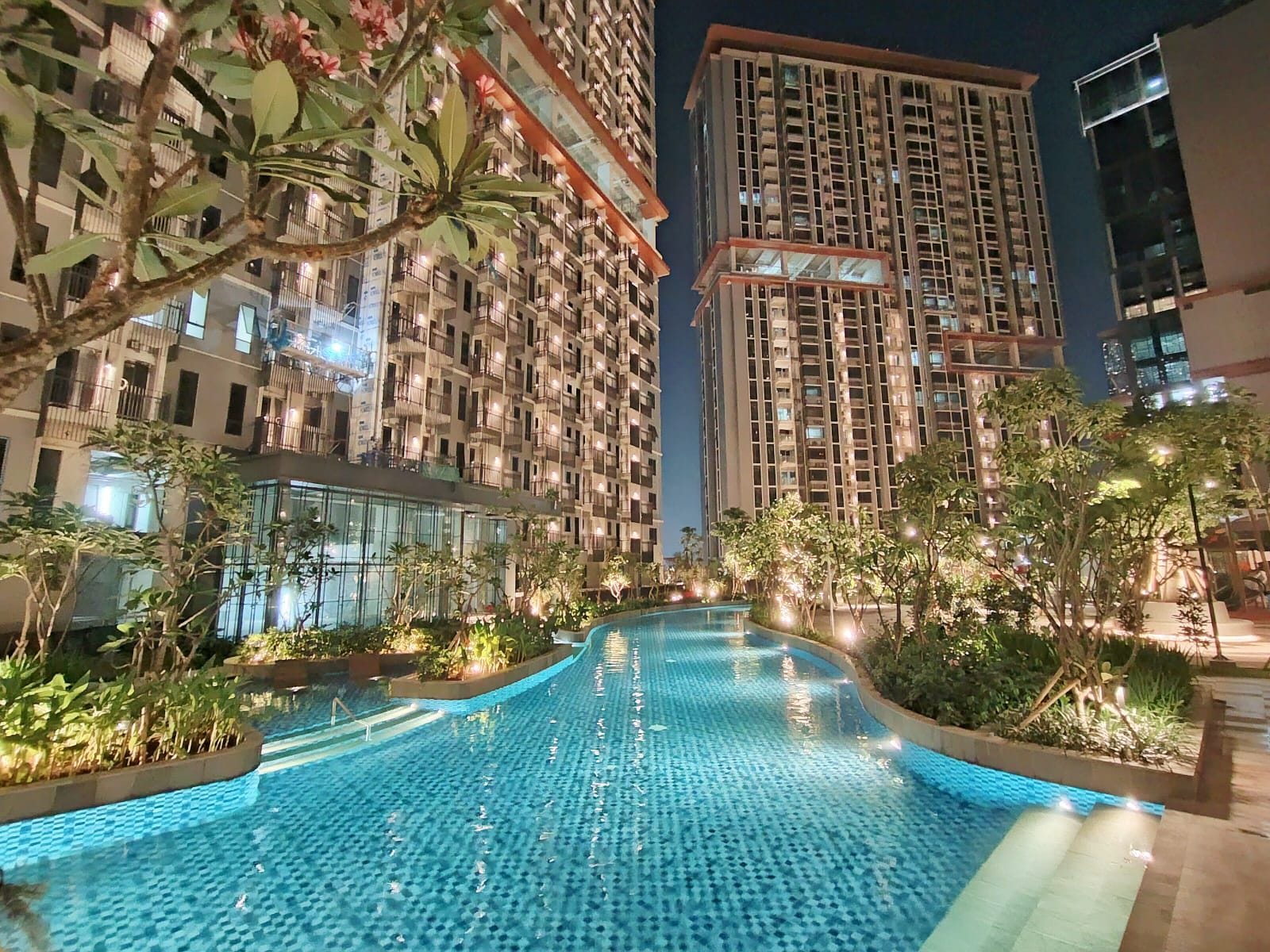Uma piscina com borda azulejada, cercada por vegetação e edifícios modernos altos à noite.