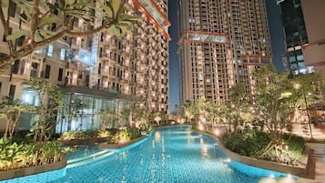 Uma piscina com borda azulejada, cercada por vegetação e edifícios modernos altos à noite.