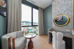Quarto de hotel moderno com uma janela grande, um espelho redondo, um sofá, uma mesinha e uma escrivaninha embutida com cadeira.