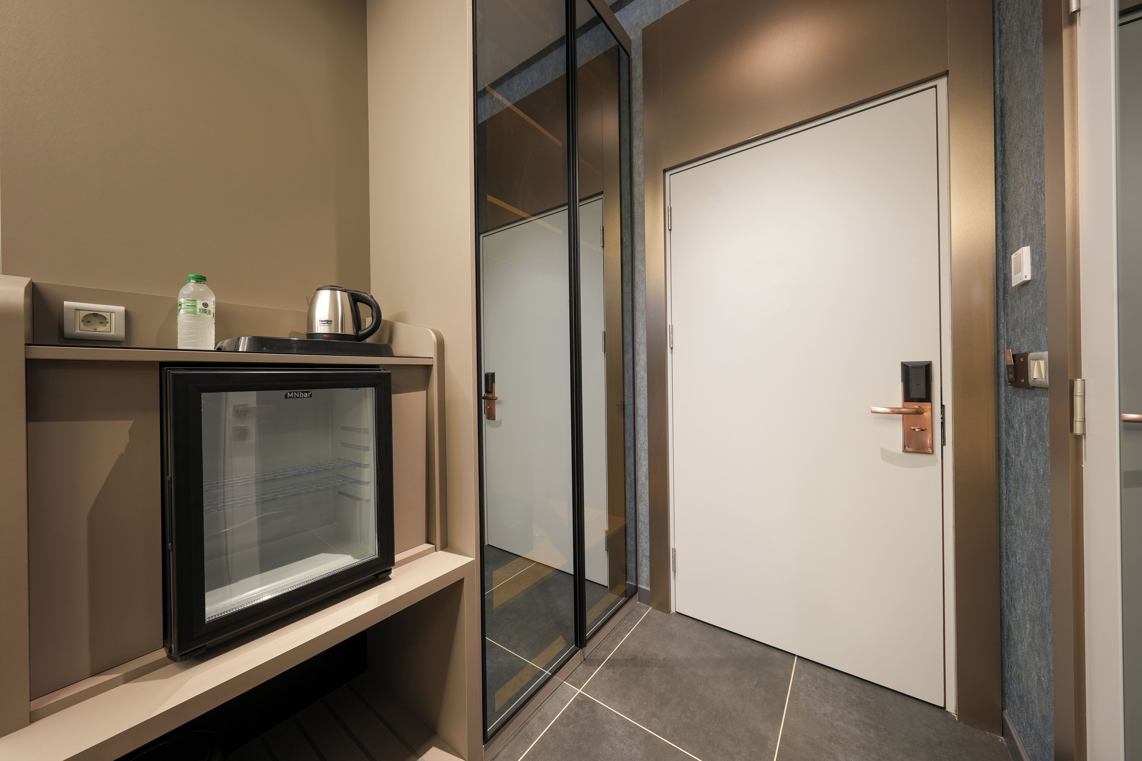 Quarto de hotel com minibar, porta de vidro para um armário e porta com puxador moderno.