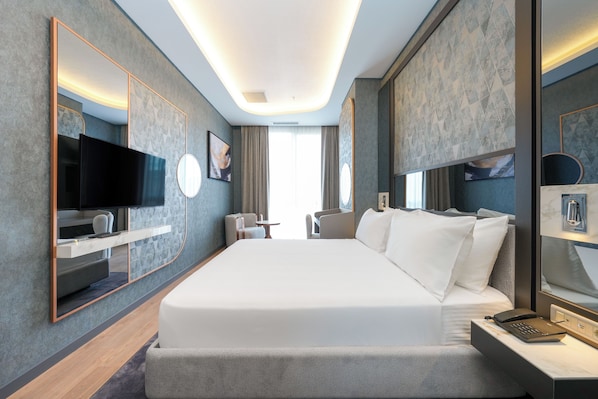Quarto de hotel moderno com uma cama grande, televisão de tela plana, escrivaninha com telefone e área de estar com poltrona e mesa.