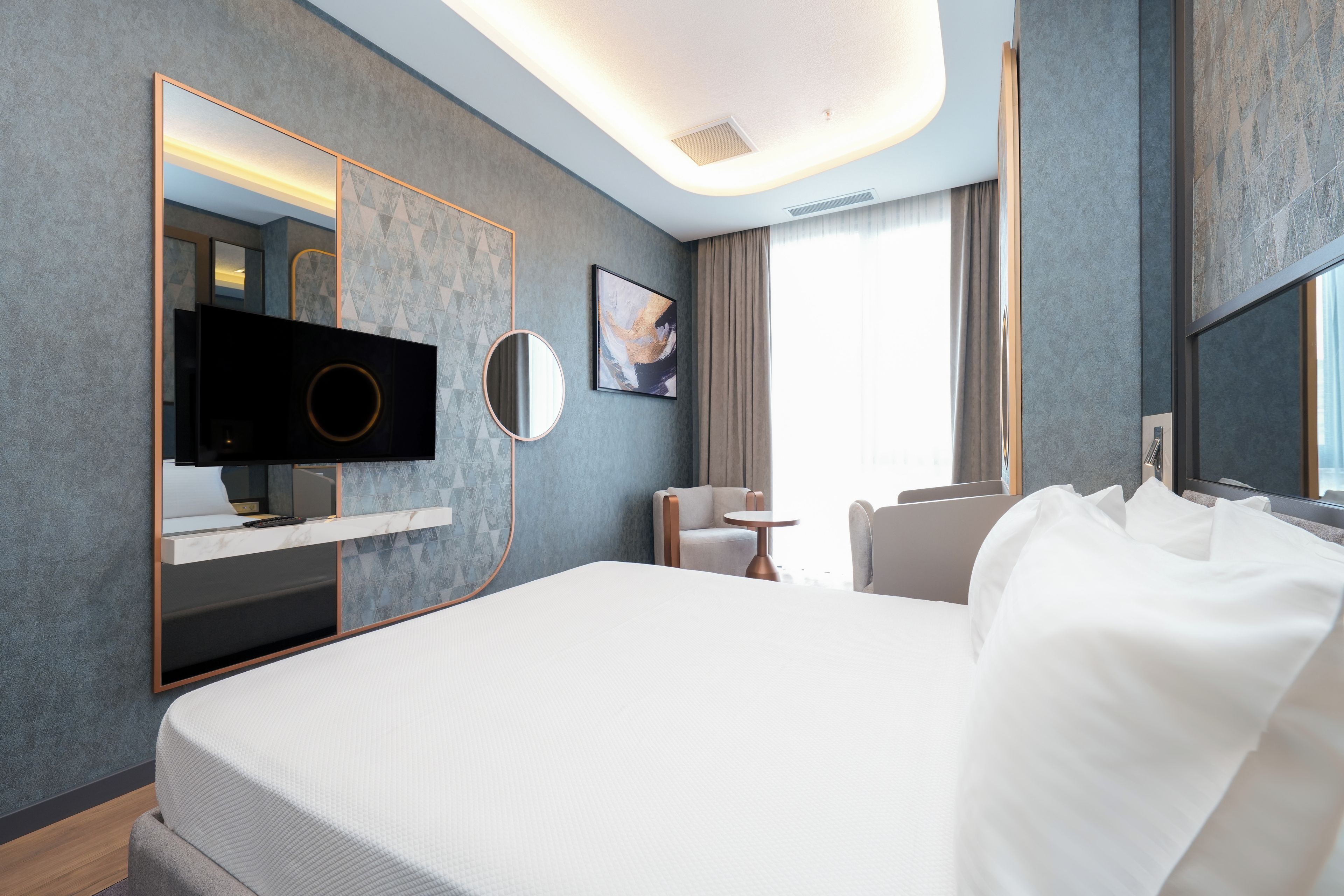 Quarto de hotel moderno com uma cama grande, televisão de tela plana, espelho redondo, mesa pequena e um sofá.