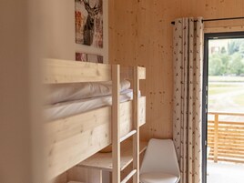 Uma cama beliche com colchão branco e uma cadeira em quarto com paredes de madeira e janela com cortinas estampadas.