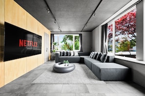 Uma sala de estar moderna com sofá seccional, TV de tela plana presa a uma parede de madeira e janelas amplas com vista para árvores.