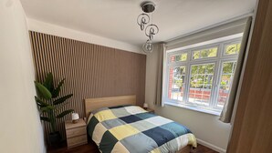 Quarto com parede de ripas de madeira, cama com colcha xadrez, mesa de cabeceira, planta em vaso e janela com vista para o exterior.
