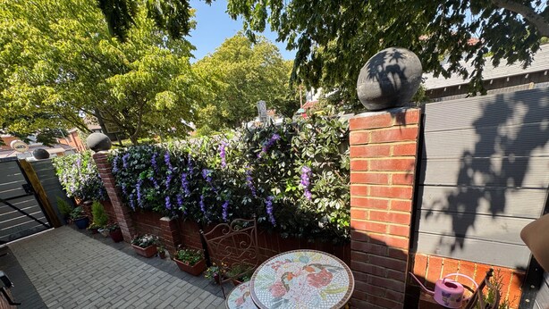 Pátio com mesa e cadeiras de azulejos, cercado por um muro de tijolos com plantas trepadeiras e um portão de metal.
