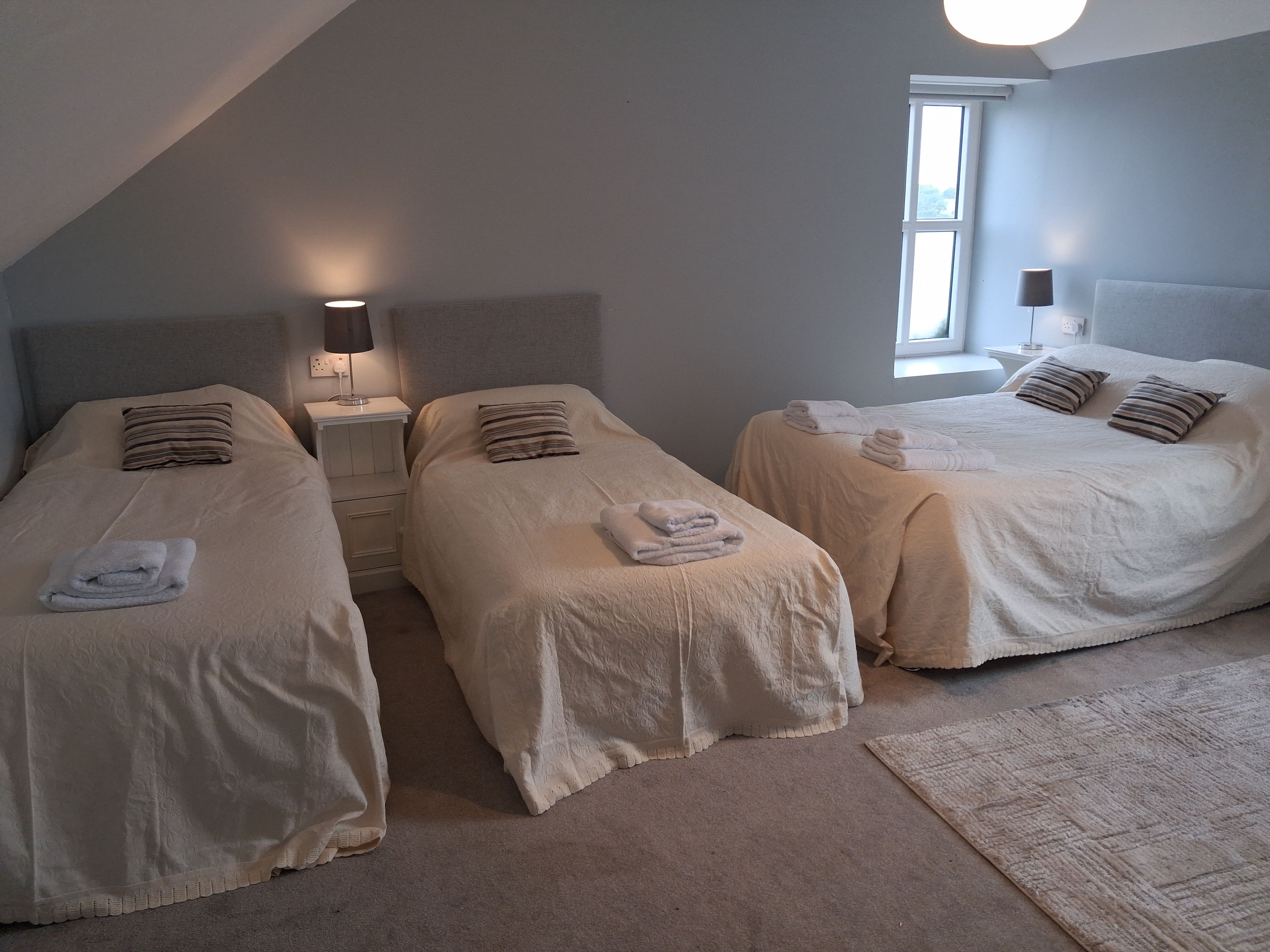 Quarto com três camas, cada uma com travesseiro e toalha dobrada. Há uma mesinha de cabeceira com abajur, uma janela com luz natural e piso coberto por carpete.
