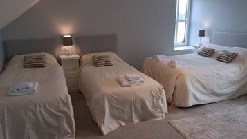 Quarto com três camas, cada uma com travesseiro e toalha dobrada. Há uma mesinha de cabeceira com abajur, uma janela com luz natural e piso coberto por carpete.