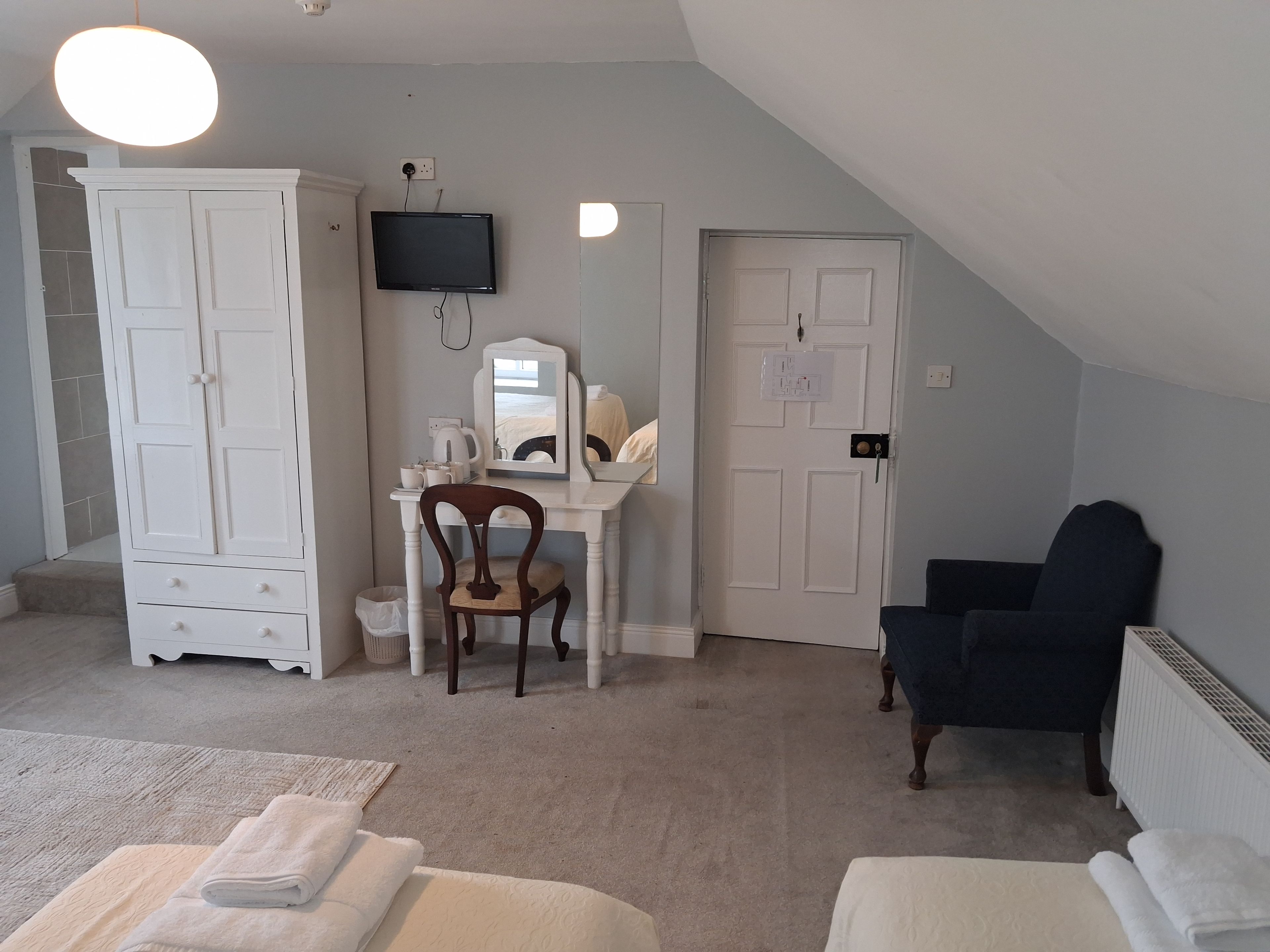 Quarto com escrivaninha branca, cadeira, guarda-roupa, televisão e porta.