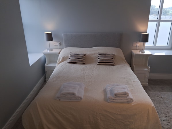 Cama bem arrumada com travesseiros e toalhas dobradas, mesa de cabeceira com abajur, e janela com vista.
