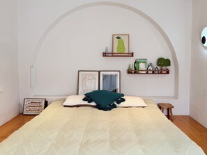 Quarto com cama, manta verde, obras de arte emolduradas e uma estante com livros e objetos decorativos.