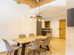 Uma área de refeições moderna com uma mesa e cadeiras de madeira, um lustre pendurado e uma área de cozinha ao fundo.